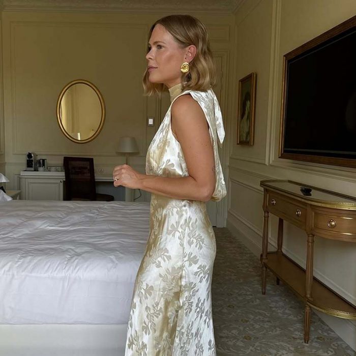 A woman in an elegant, floral-patterned dress stands in a luxurious bedroom with cream-colored walls, a round mirror, and classic furniture.