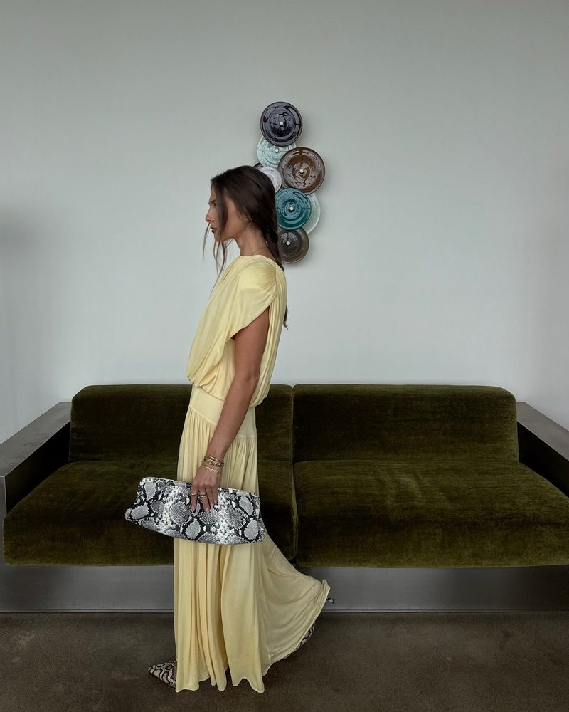 A woman in a flowing yellow dress stands in profile%2C holding a snakeskin-patterned clutch. Behind her is a dark green sofa and decorative wall plates. The scene conveys elegance and calm.