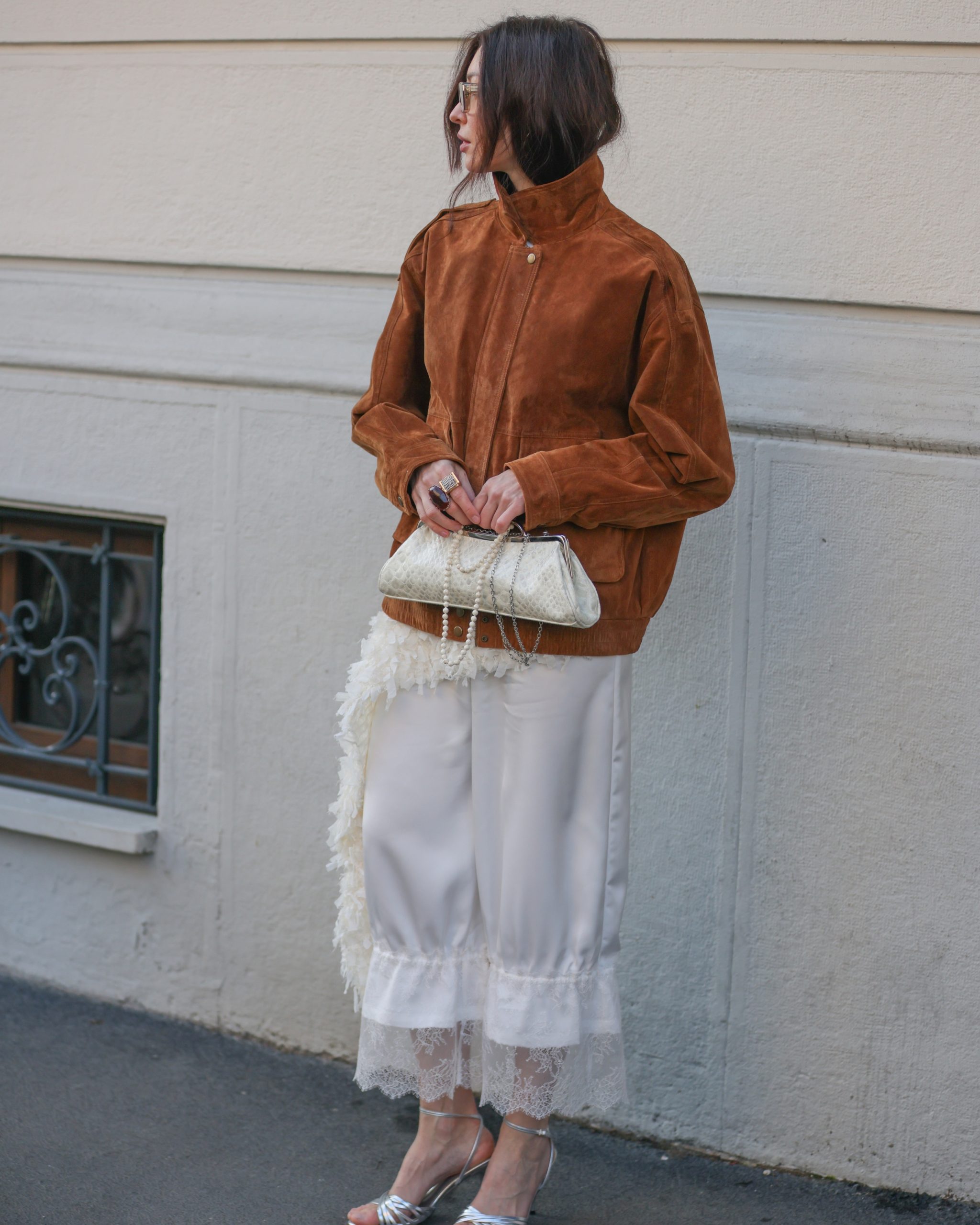 Suede brown jacket paired with a cream satin pleated skirt featuring lace trim%2C complemented by silver strappy heels and a metallic clutch
