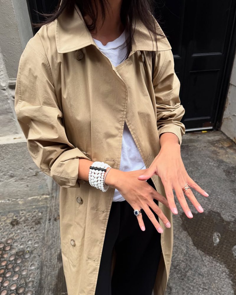 Beige trench coat layered over white tee with black skirt%2C styled with statement bracelets and rings for chic minimalist outfit