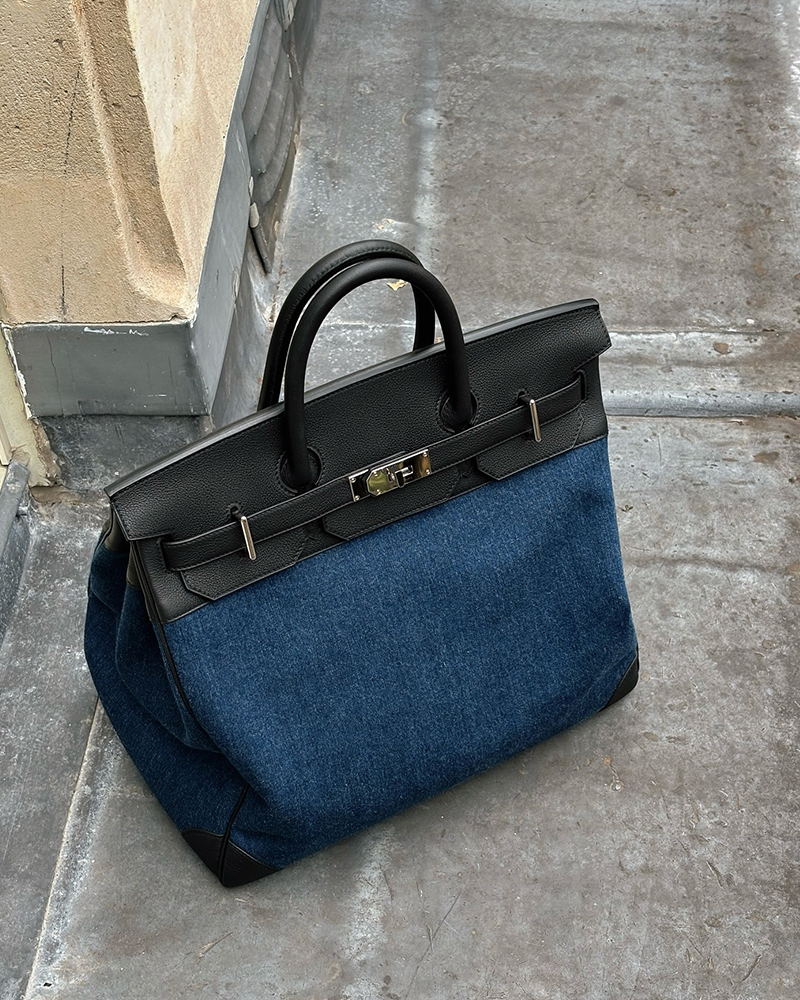 Herm%C3%A8s Birkin bag in blue denim with black leather trim and top handles%2C placed on concrete pavement