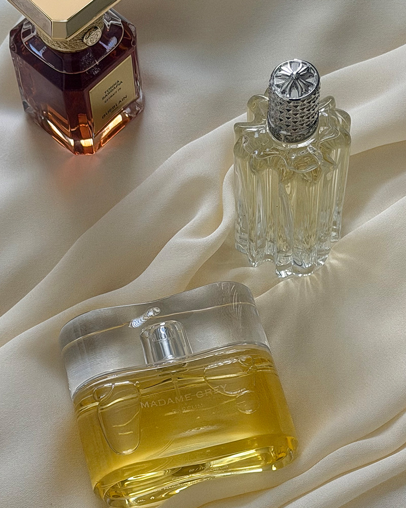 Luxury perfume bottles including Guerlain and Chanel fragrances arranged on soft ivory fabric with warm lighting backdrop