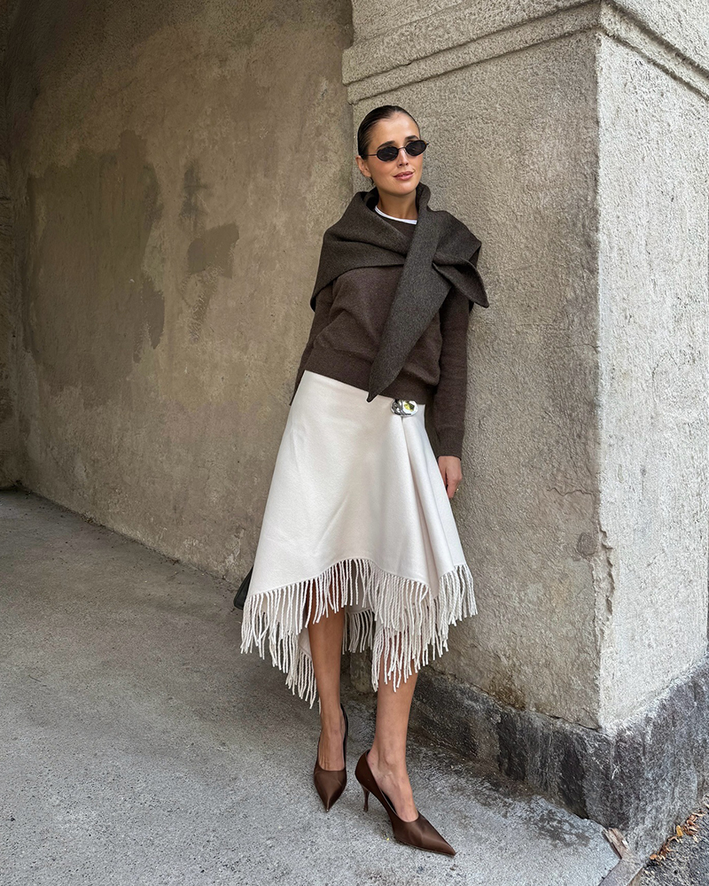 Woman in brown draped knit sweater%2C cream asymmetrical fringe midi skirt and brown pointed heels with sunglasses%2C posed on a city sidewalk