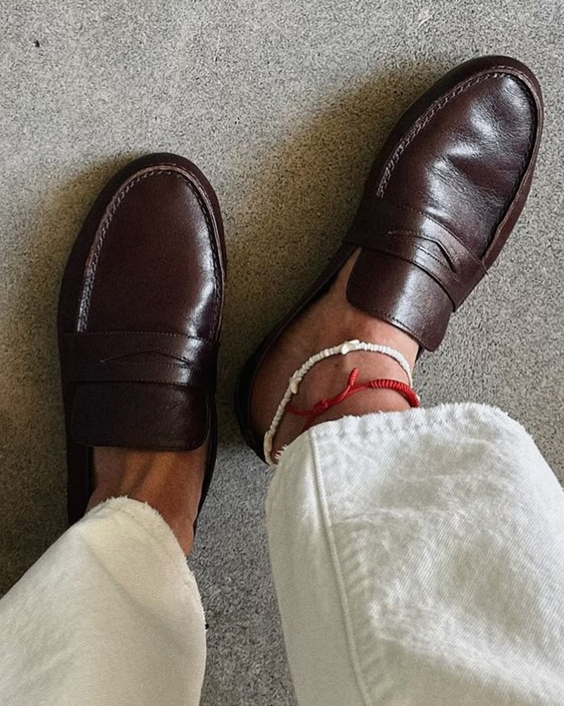 Brown leather penny loafers styled with cream straight-leg trousers and ankle bracelets on concrete floor