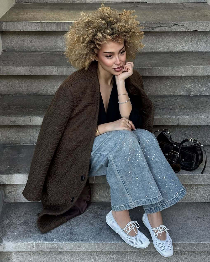 Model wearing a brown textured coat over a black top%2C embellished wide-leg jeans%2C and white ballet sneakers%2C sitting on stone steps.