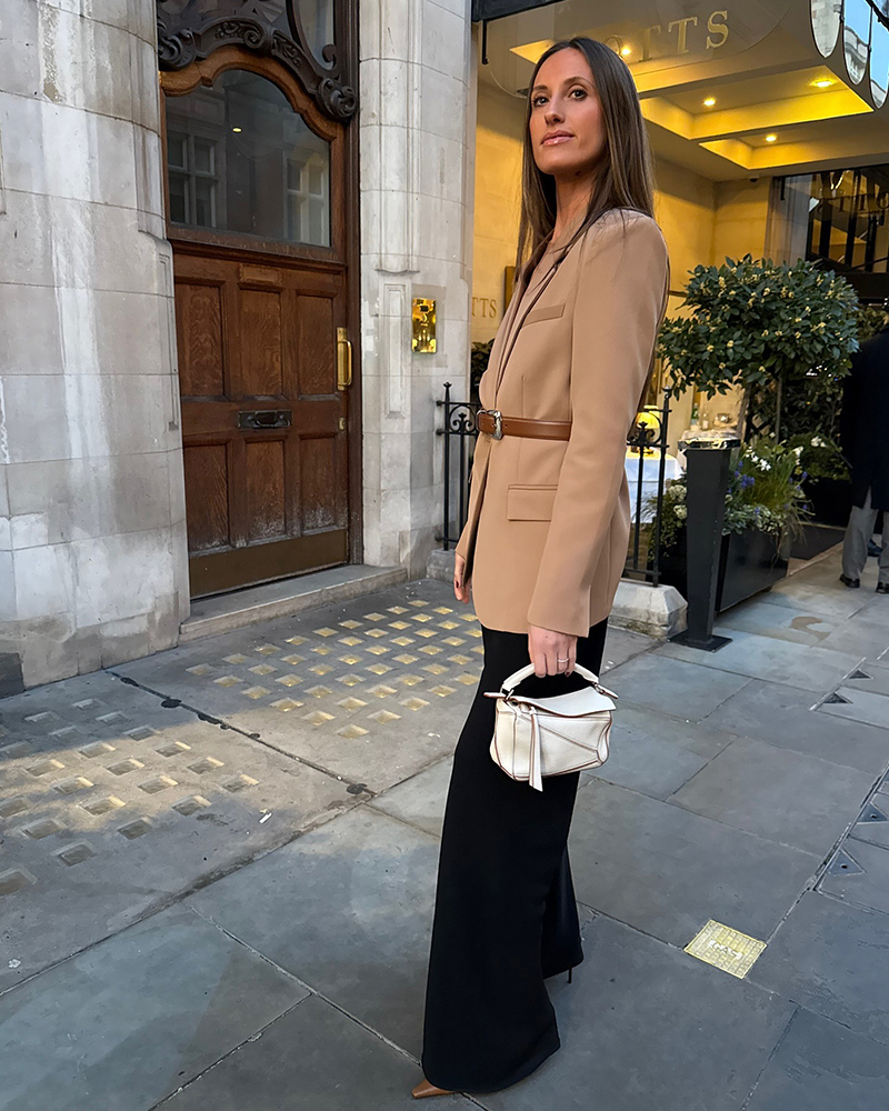 Woman in tailored beige belted blazer with black wide-leg trousers and white mini top-handle bag%2C standing on city sidewalk outside boutique