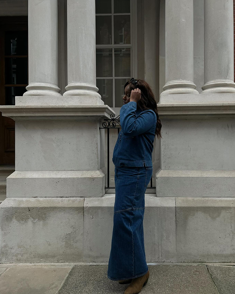 Woman in dark wash denim jacket and long denim skirt with ankle boots%2C styled on city street against stone building facade