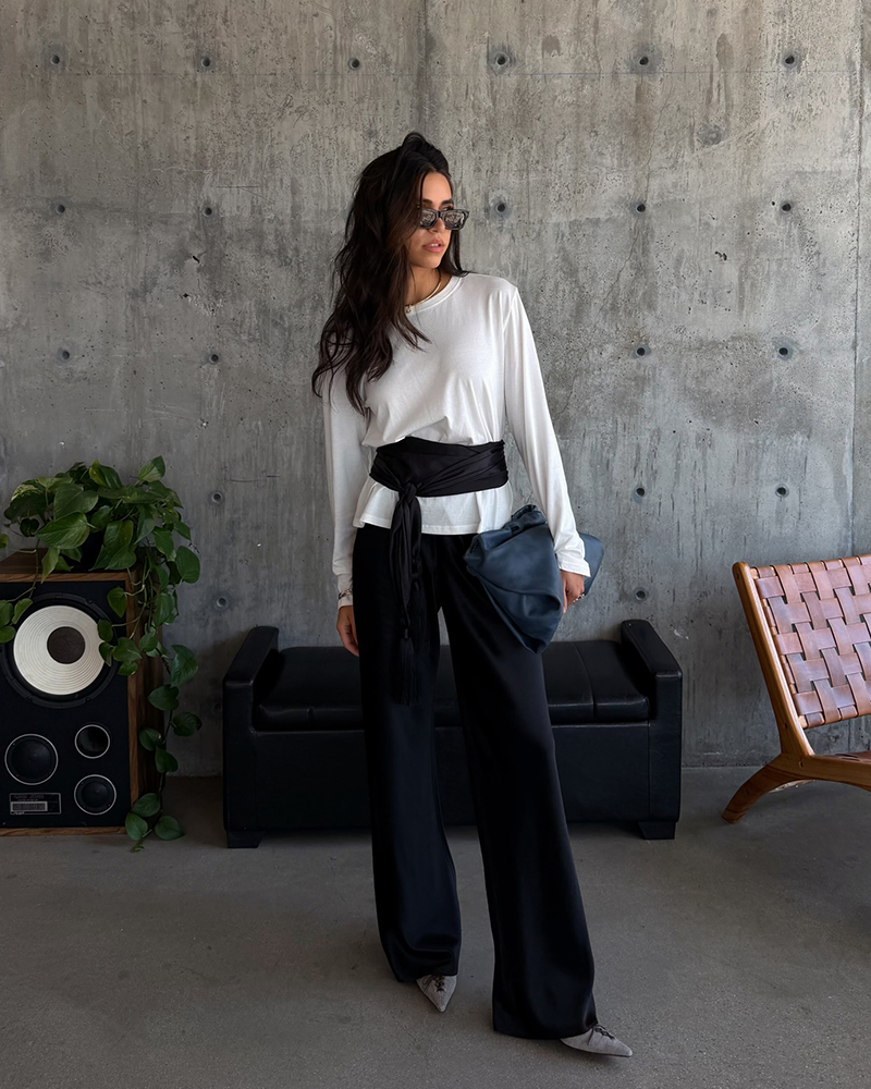 Woman in white long-sleeve top with black wide-leg flared pants and wrap belt%2C holding clutch in modern minimalist interior