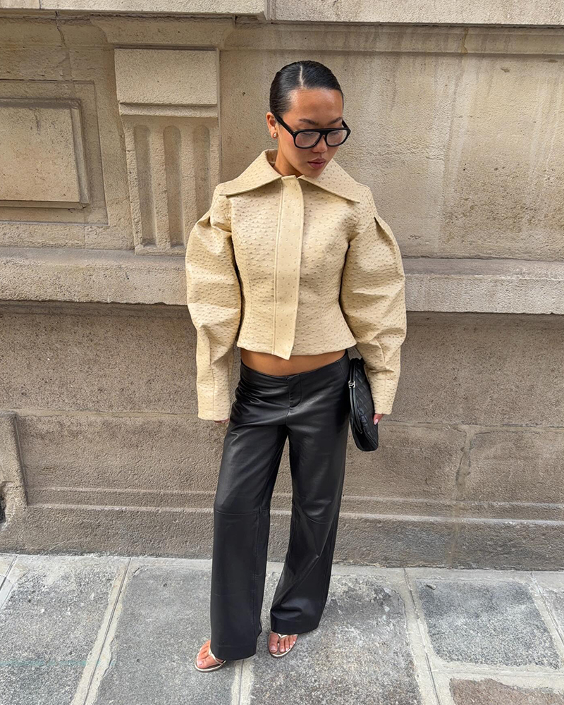 Woman in beige textured cropped jacket with puff sleeves%2C black leather wide-leg pants%2C glasses and sandals%2C styled on a city sidewalk backdrop
