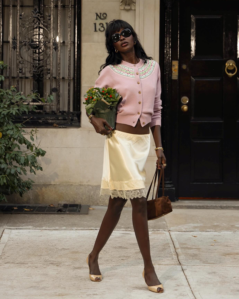 Woman in pink embroidered cardigan with silk slip skirt%2C sunglasses%2C brown mini bag and heels%2C walking on city street with flowers