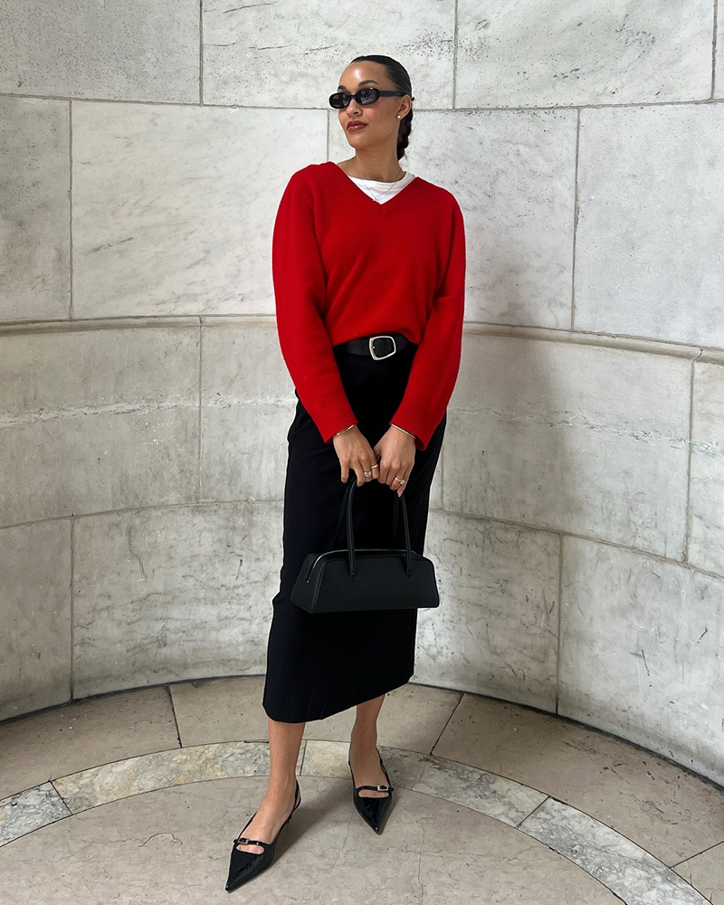 Woman in red knit sweater with black midi skirt%2C belt%2C pointed heels and top-handle bag%2C styled against stone wall backdrop