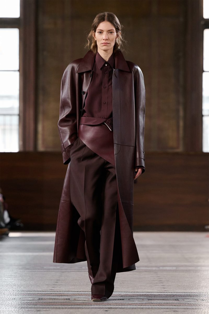 Runway model in burgundy leather trench coat with matching shirt and tailored trousers, walking on minimalist indoor fashion runway
