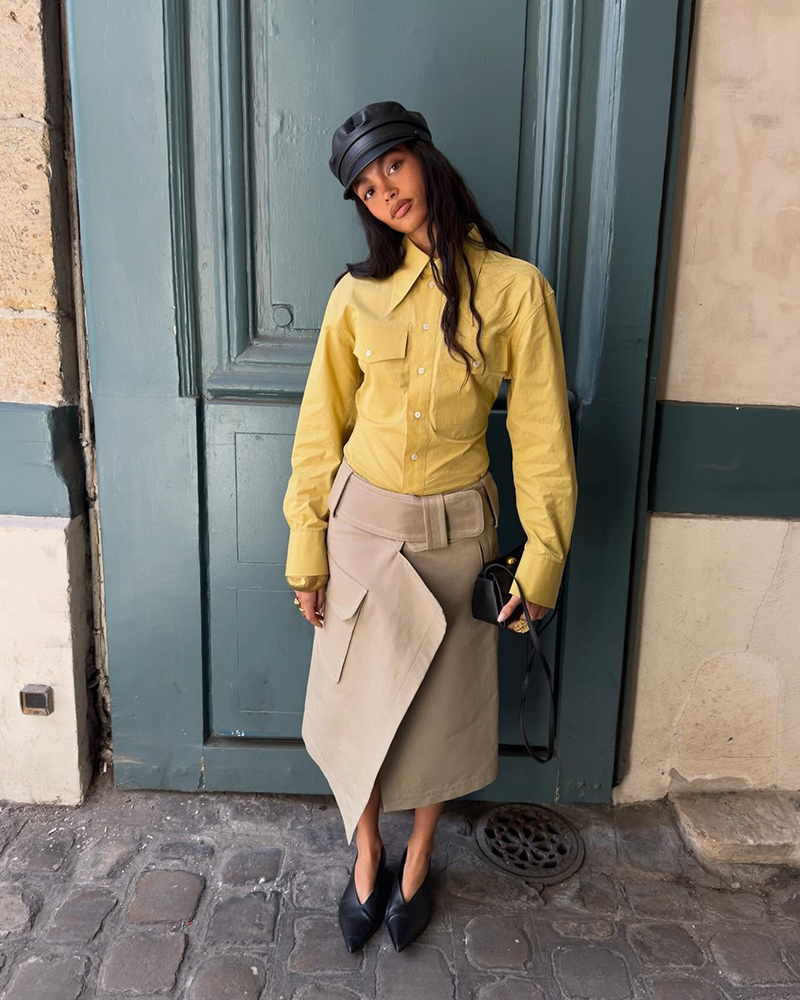 Woman in yellow button-down shirt with beige wrap midi skirt%2C black heels and leather cap%2C standing by green doorway on cobblestone street