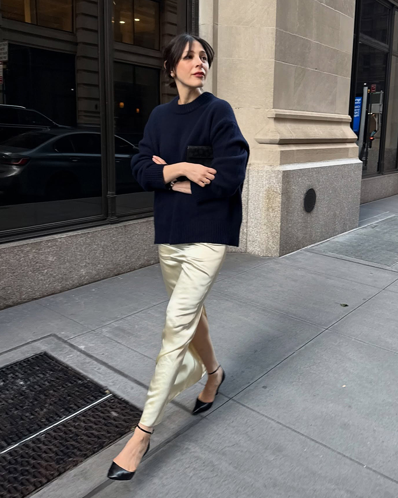 Woman in navy crewneck sweater with satin midi skirt and black pointed heels%2C walking on city sidewalk in minimalist spring outfit