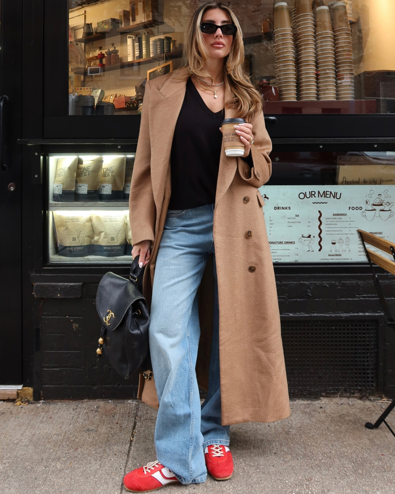 Street style look with camel coat%2C black top%2C wide-leg jeans%2C red sneakers and leather backpack outside caf%C3%A9 setting