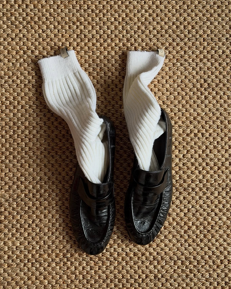 Black leather penny loafers paired with white ribbed crew socks%2C styled on woven jute rug for casual transitional outfit