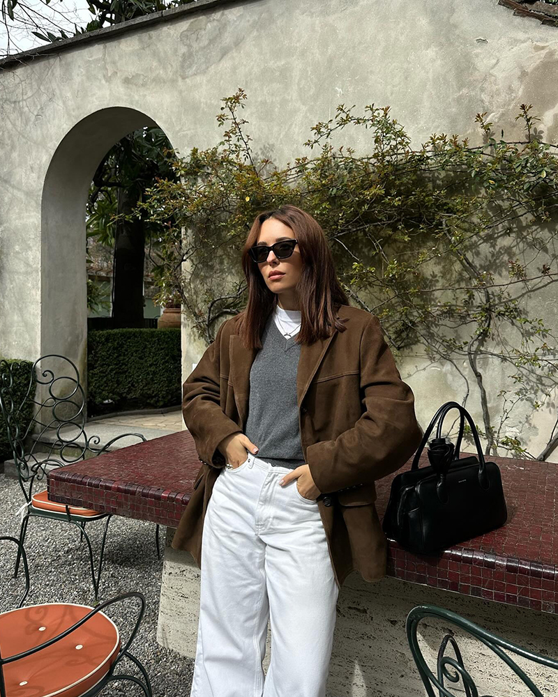 Woman in brown suede jacket%2C gray knit sweater and white jeans with black handbag%2C styled in outdoor courtyard setting