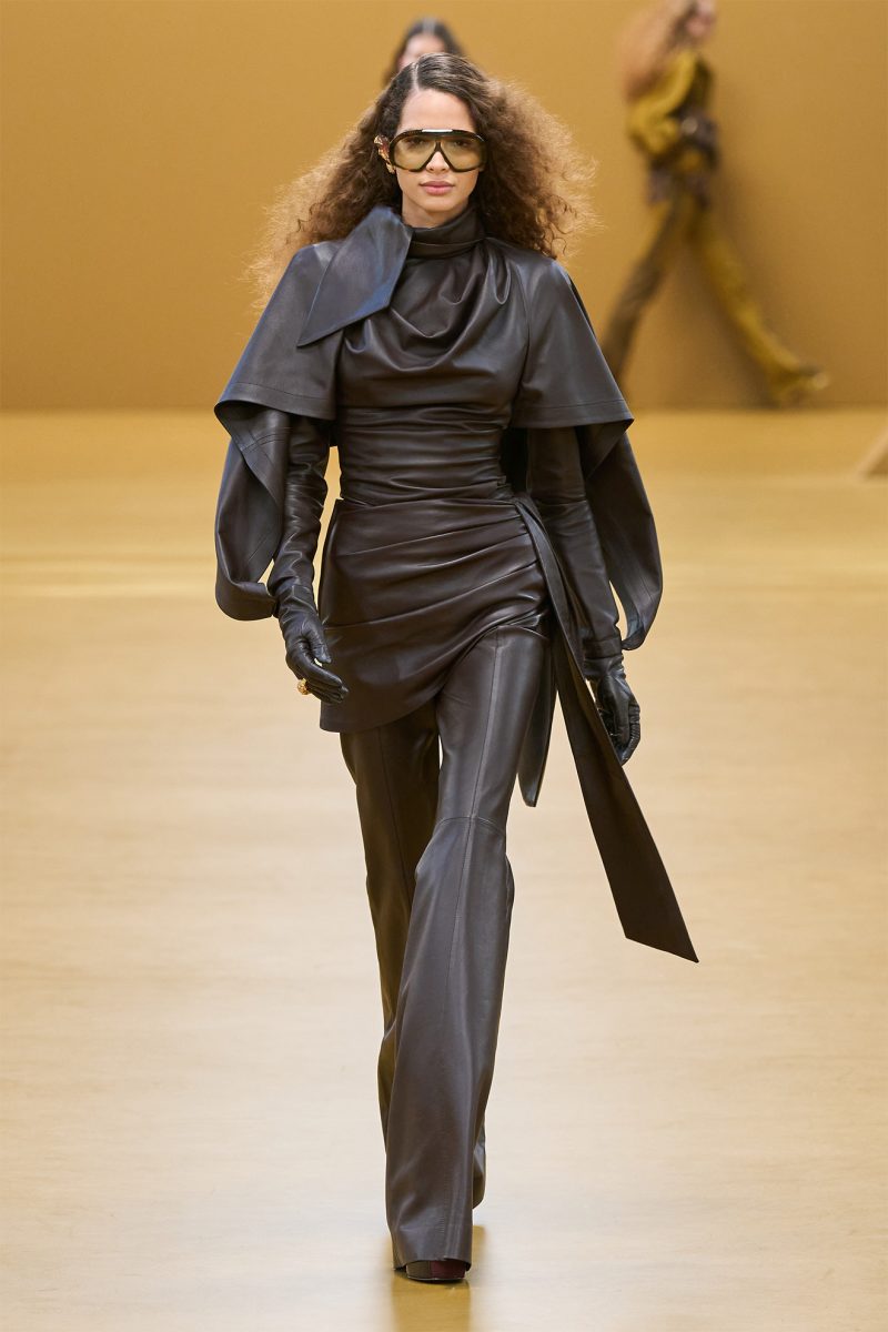 Runway model in black leather draped top and matching trousers with gloves and oversized sunglasses on minimalist runway backdrop