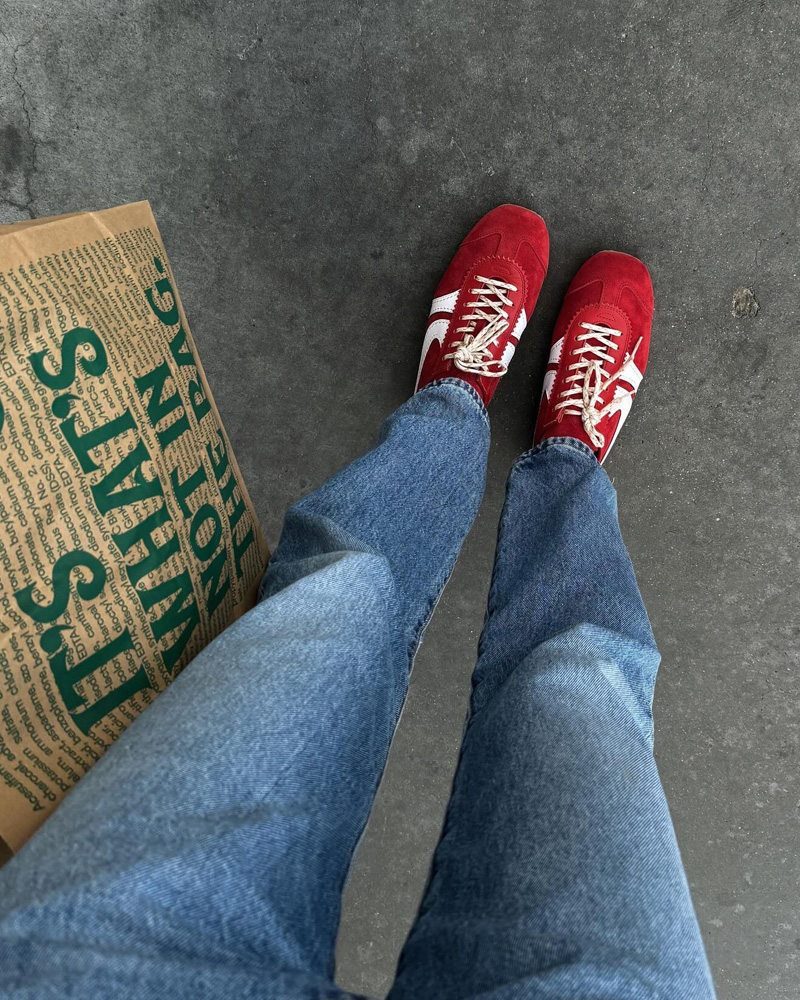 Top-down view of red suede sneakers with white laces styled with blue jeans on concrete floor beside shopping bag