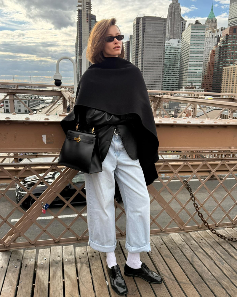 Street style featuring cuffed light-wash jeans with black cape coat%2C leather handbag%2C loafers and sunglasses on a city bridge backdrop