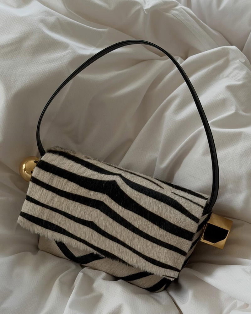 Zebra-print calf hair mini shoulder bag with black leather strap and gold hardware%2C styled on soft white bedding