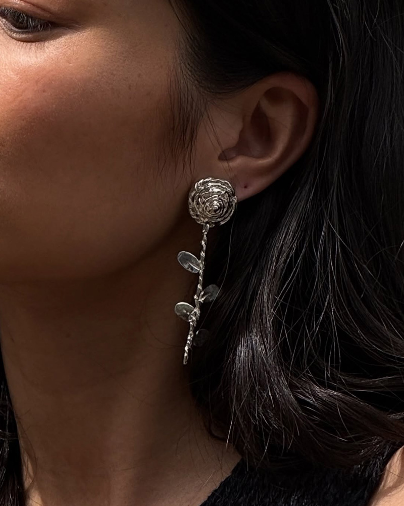Close-up of sculptural silver floral drop earring with textured petals%2C worn with sleek hair against a minimal backdrop