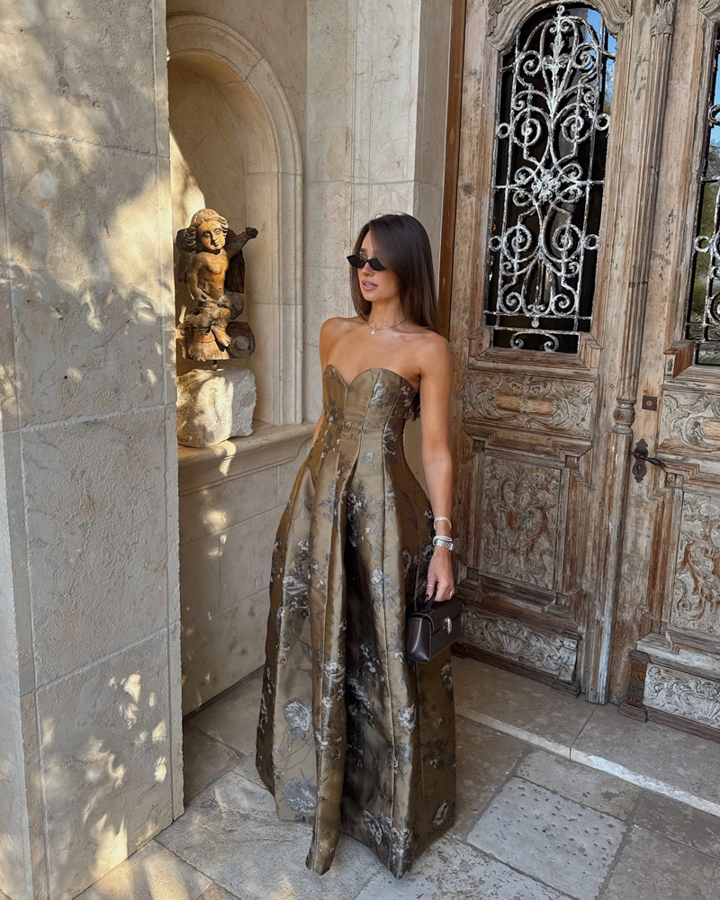 Woman in strapless bronze floral evening gown with sunglasses and mini handbag standing by ornate wooden doors in sunlit courtyard
