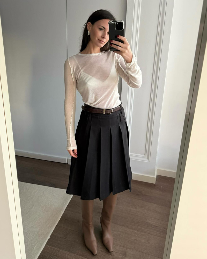 Mirror selfie outfit with sheer long-sleeve top%2C pleated black midi skirt%2C leather belt and knee-high boots in a minimal interior