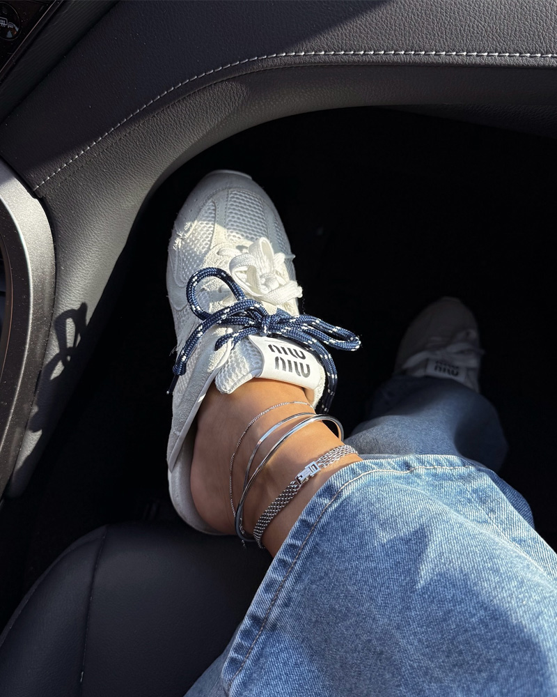 Close-up of Miu Miu white sneakers with blue laces and silver ankle bracelets%2C styled with light-wash denim inside car interior