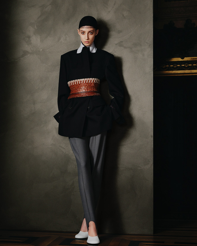 Model in structured black blazer with statement corset belt%2C tailored gray trousers%2C and white flats against moody textured backdrop