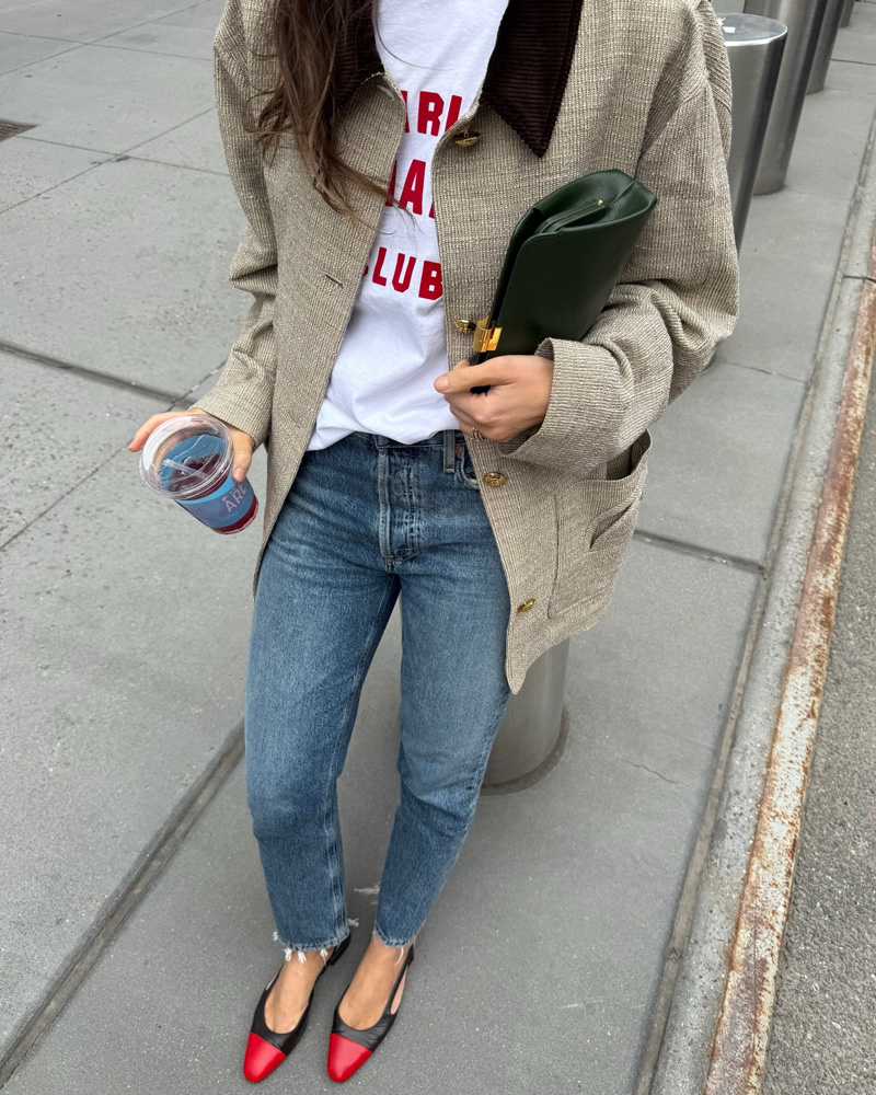 Casual street style outfit with beige textured jacket%2C graphic tee%2C blue jeans%2C red cap-toe flats%2C and green clutch on city sidewalk