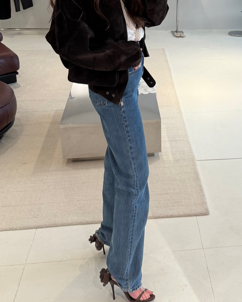 Casual chic outfit with dark suede jacket%2C white blouse%2C straight-leg blue jeans and strappy heeled sandals in a minimalist boutique setting