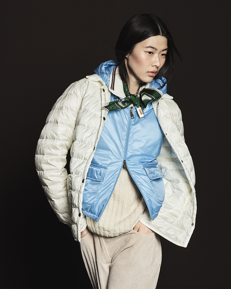 Moncler layered outfit with a white quilted jacket%2C blue hooded vest and green silk scarf against a dark studio backdrop