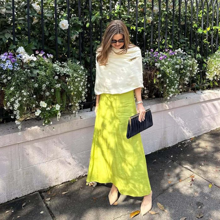A woman wearing sunglasses, a white top, and a bright yellow skirt stands on a sidewalk in front of a flower-filled garden bed and black metal fence, holding a black clutch.
