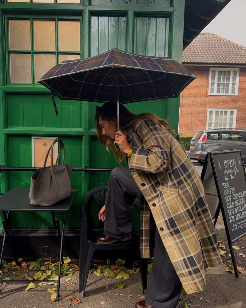Chic plaid trench coat paired with wide-leg trousers%2C holding a black umbrella while a stylish gray tote rests on a cafe table