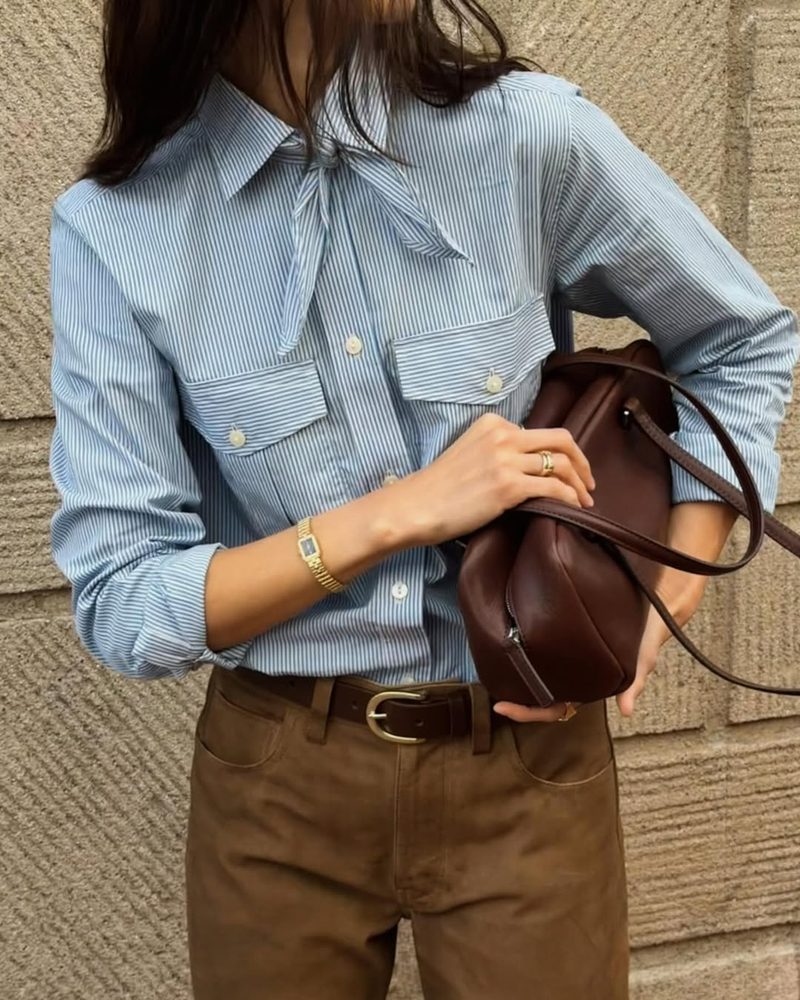 Blue striped button-down shirt with tie-neck detail paired with brown trousers and leather shoulder bag for polished everyday style