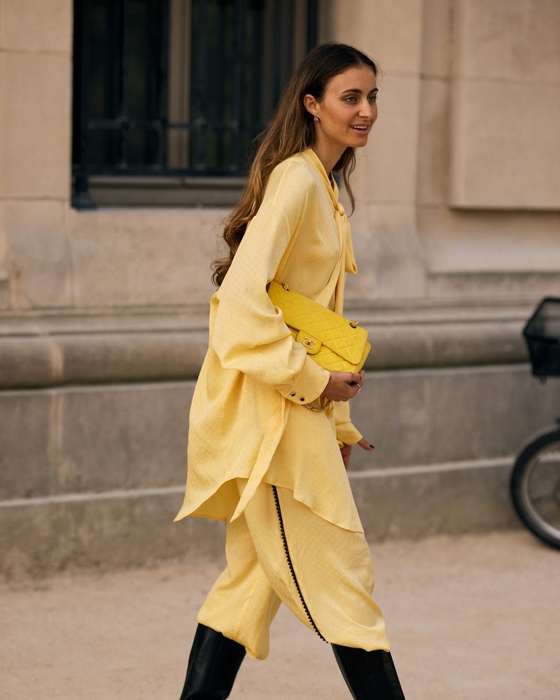 Model wearing a flowing yellow silk blouse and matching pants with black side trim%2C paired with black leather knee-high boots and a yellow quilted Chanel handbag.