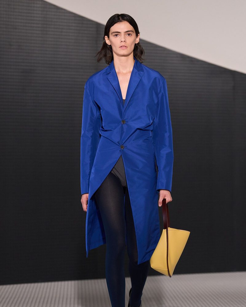 Jil Sander spring collection showcases a vibrant blue trench coat paired with sleek black tights and modern heeled shoes%2C complemented by a minimalist yellow tote bag