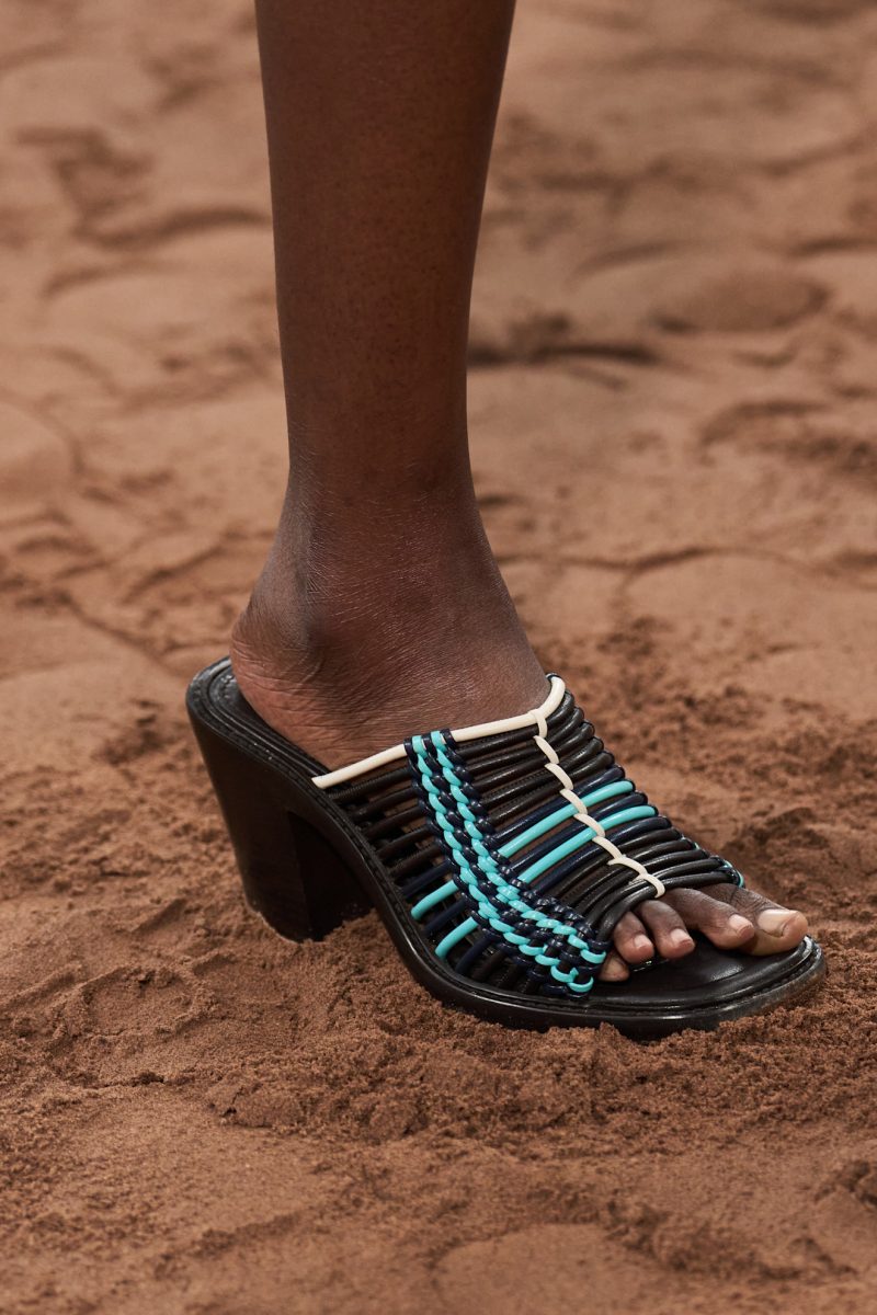 Burberry black heeled mule sandal with woven blue and white straps on sandy ground