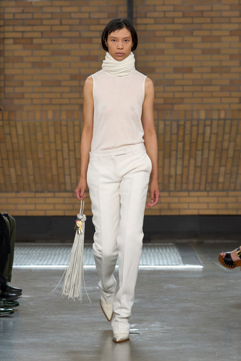 Model wearing Calvin Klein sleeveless white turtleneck top with tailored white trousers and carrying a white fringe handbag in a minimalist runway setting