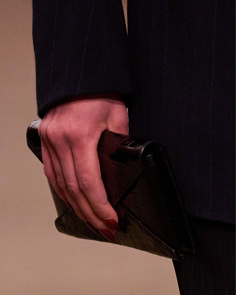Close-up of Saint Laurent dark leather clutch held against a black pinstripe suit on the runway%0A