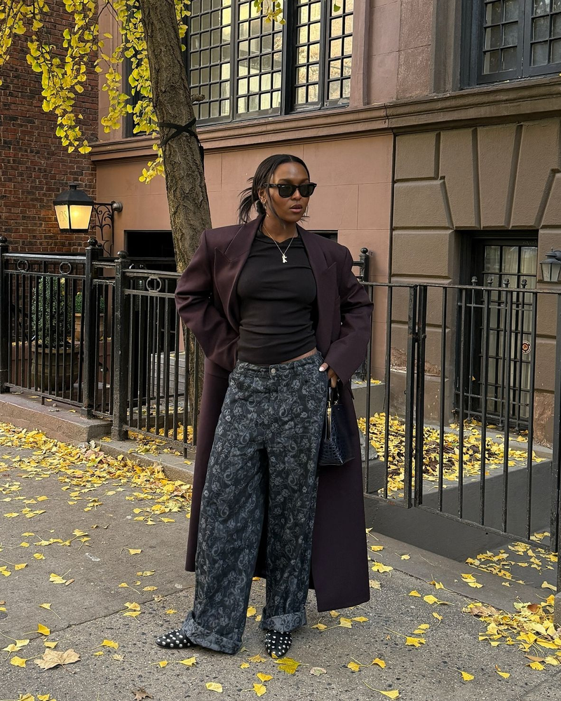 Woman wearing dark floral printed wide-leg jeans with a black top%2C long plum coat%2C studded black shoes%2C and sunglasses in autumn street style