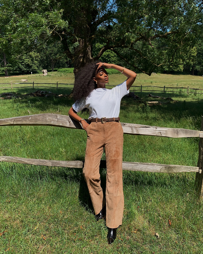 Model wearing tan suede pants with a brown belt%2C white short-sleeve top%2C and black ankle boots in a sunny outdoor setting