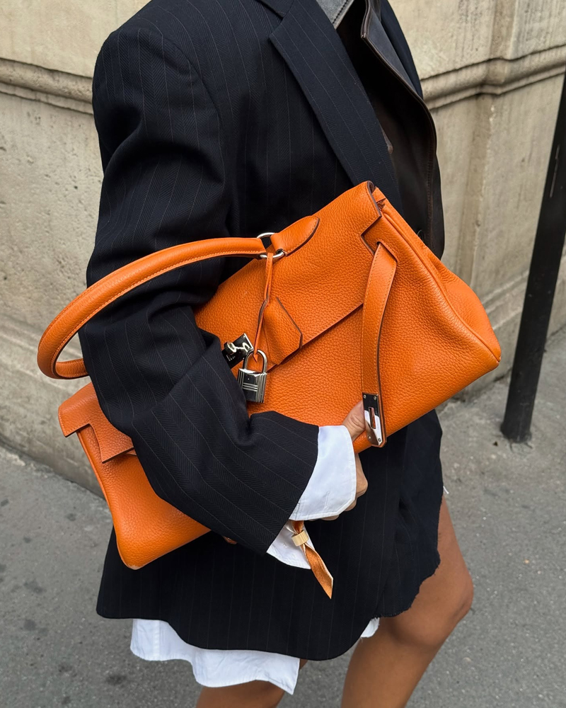 Close-up of orange pebbled leather top-handle handbag with silver lock%2C styled with black pinstripe blazer on city street