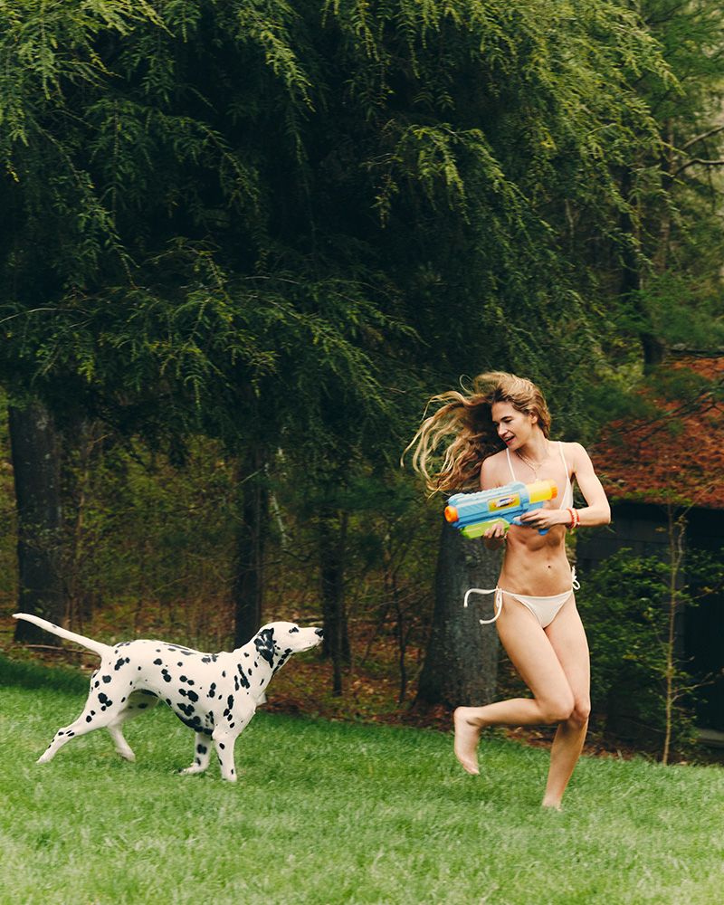 model wearing a beige string bikini playing with a dalmatian dog outdoors in a green forest setting summer swimsuit style