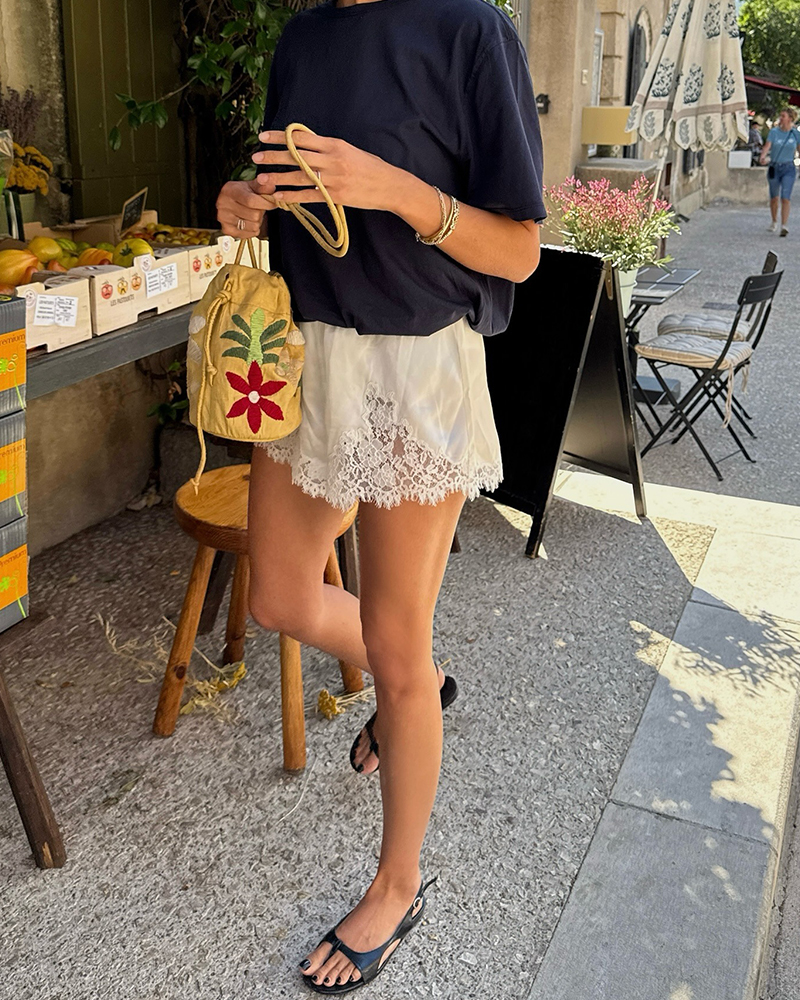Navy tee with white lace silk shorts%2C black sandals and embroidered drawstring bag styled at outdoor market cafe