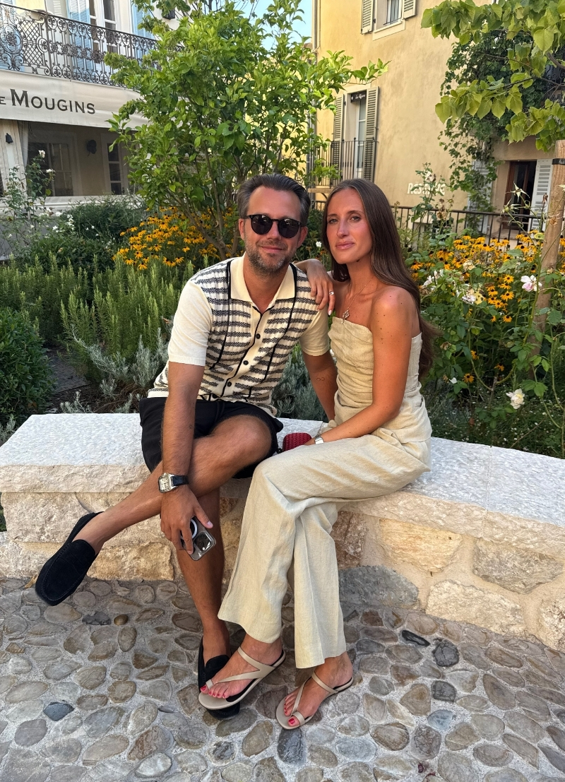 Couple in neutral summer outfits, linen jumpsuit and striped knit polo with shorts, seated in garden courtyard with flowers