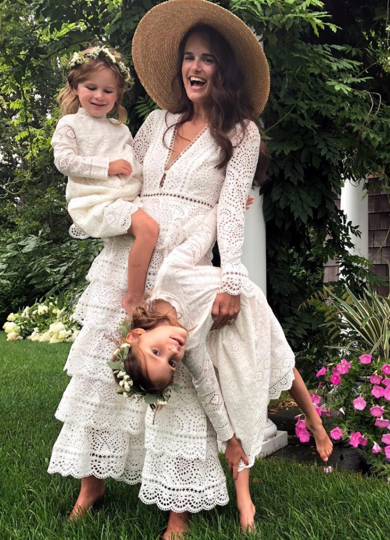 Mother in white lace dress with straw hat holding children, styled in blooming garden with soft romantic spring vibe