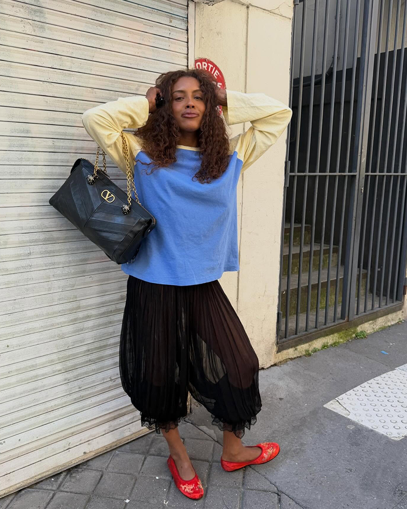 Woman in blue layered sweatshirt with black sheer balloon pants%2C red flats and chain-strap bag%2C posing on urban street