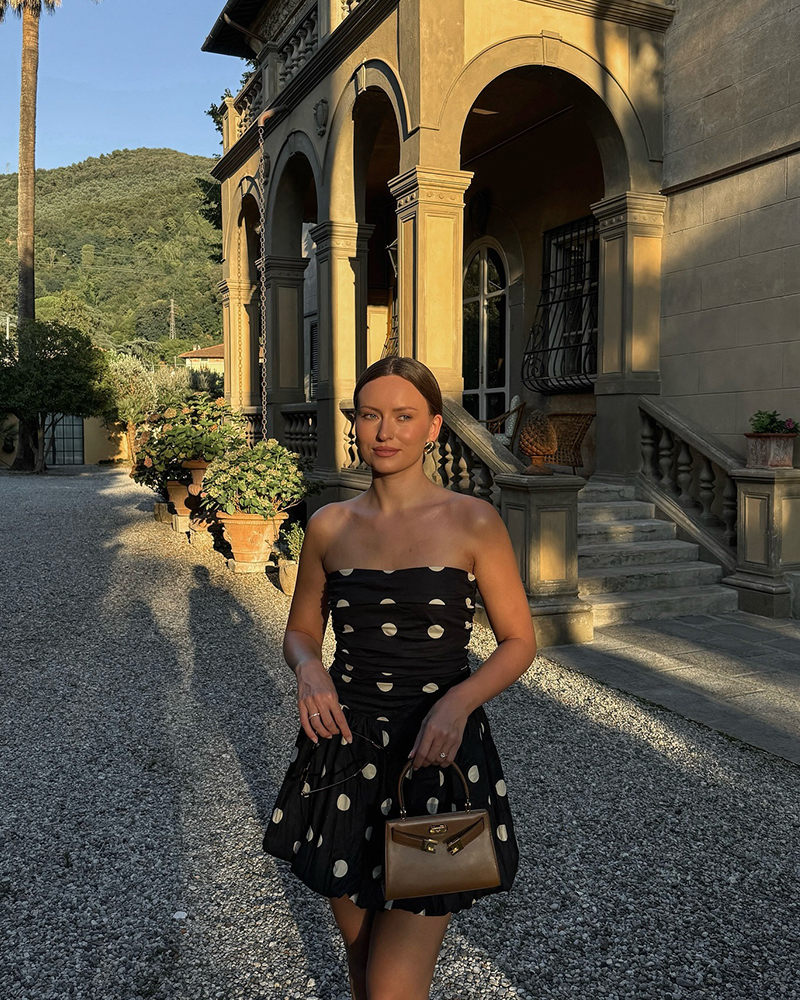 Black strapless polka dot mini dress with top-handle leather bag and sandals styled in sunlit villa courtyard